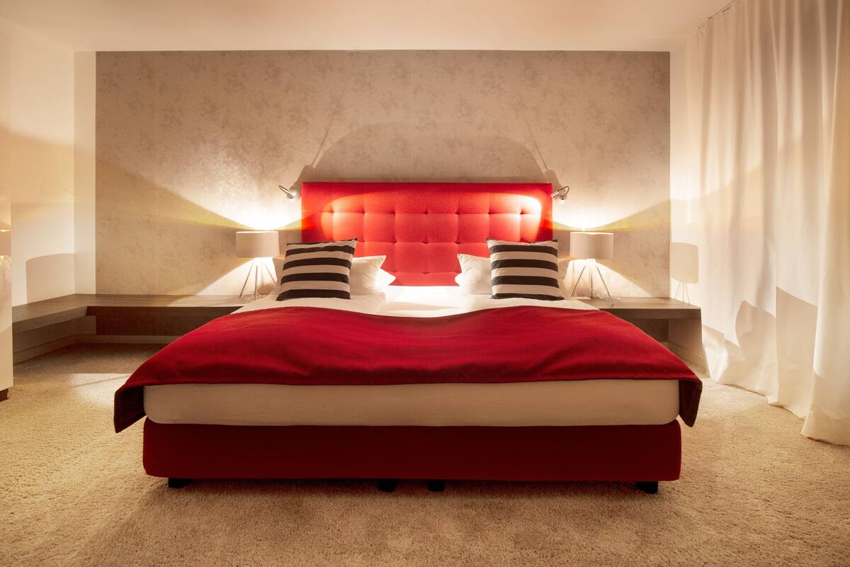 Quarto moderno com cabeceira estofada, colcha vermelha e duas luminárias de apoio.