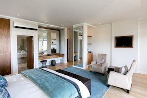Quarto de hotel moderno com uma cama grande, área de estar com uma poltrona, uma mesa pequena e uma TV fixada na parede.