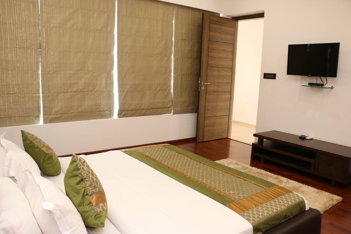 Quarto de hotel com cama, televisão, armário de madeira e uma janela grande com cortinas.
