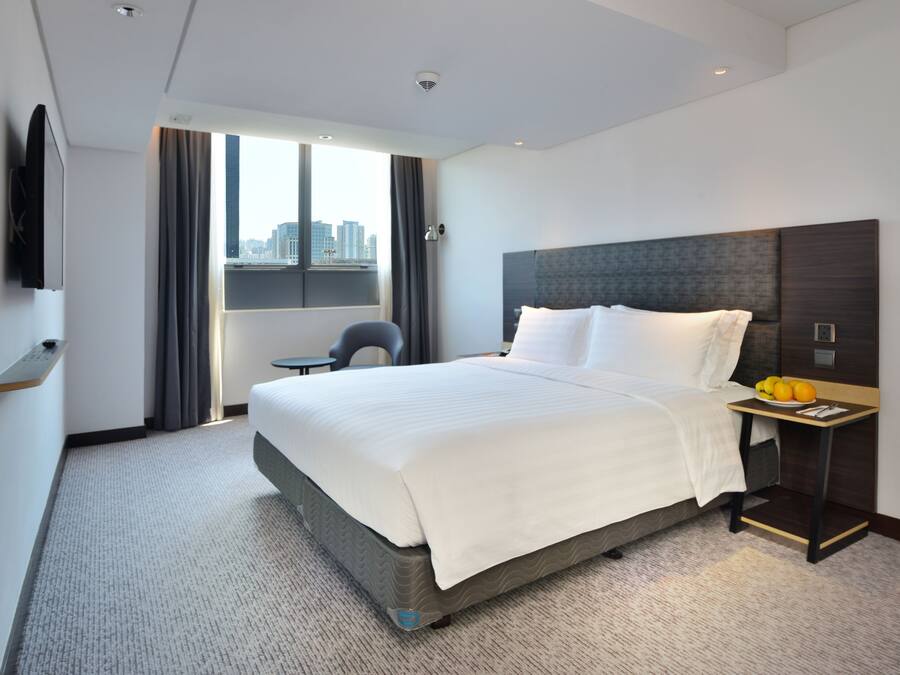 Quarto de hotel moderno com uma cama grande, uma mesinha de cabeceira com frutas, uma cadeira e vista para a cidade pela janela.