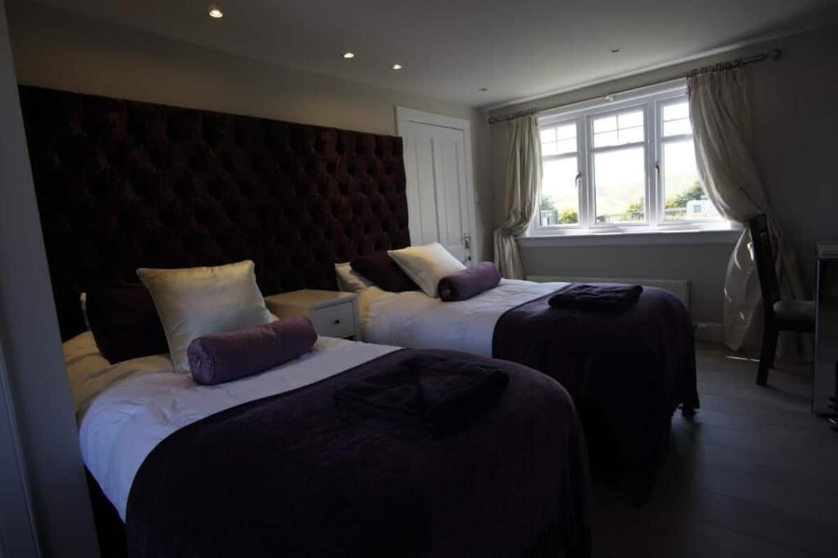 Quarto de hotel com duas camas, cabeceira escura, janela com cortinas e uma cadeira.