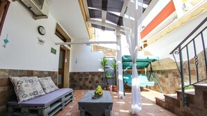 Uma área de estar coberta ao ar livre, com um banco, mesa, plantas em vasos e uma escadaria.