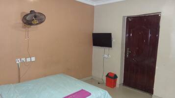 Quarto com cama, televisão na parede, porta e uma pequena lixeira.
