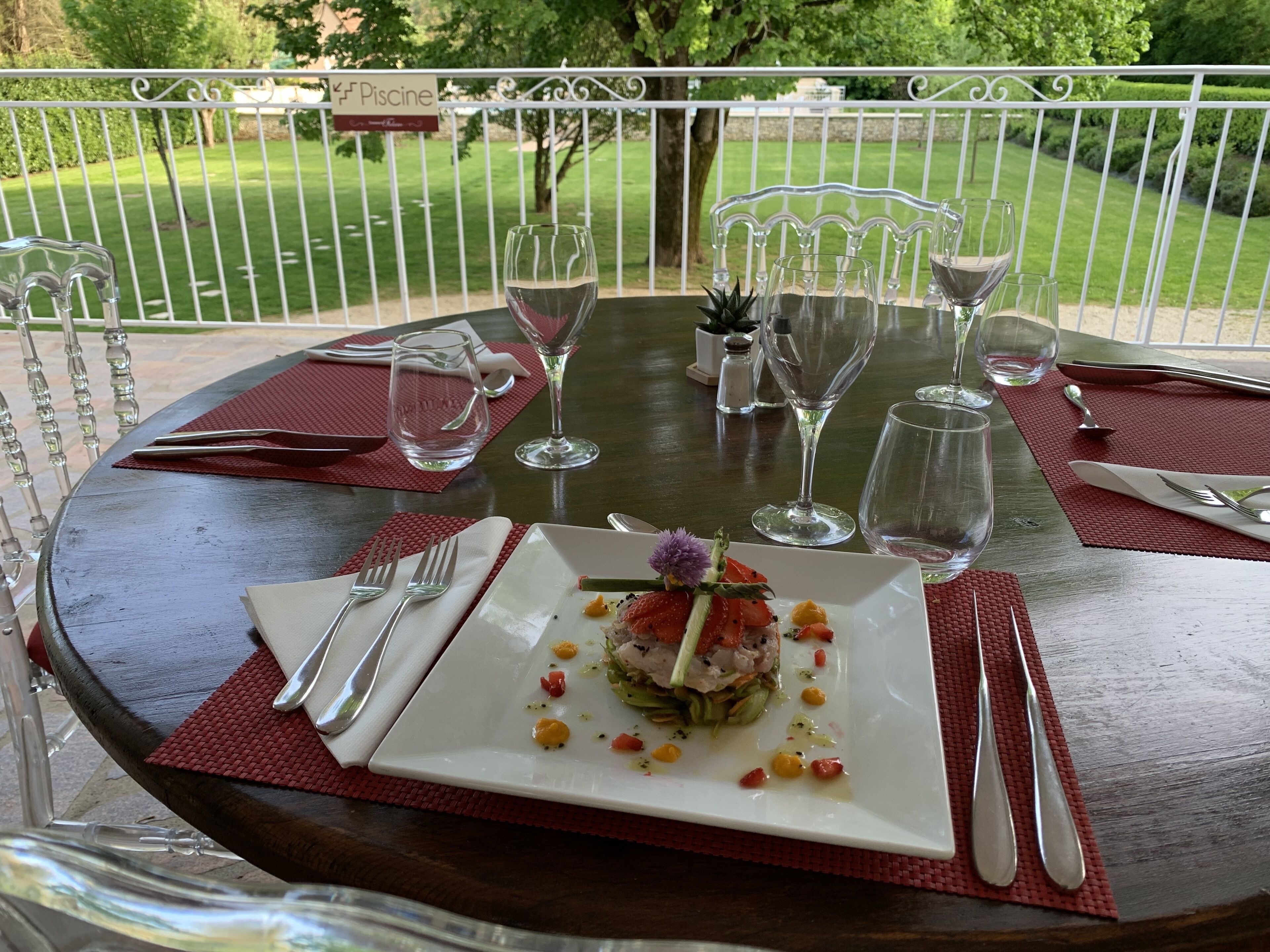 Mesa de jantar posta ao ar livre, com prato de comida, talheres e copos de vinho.