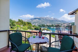Uma varanda com duas cadeiras verdes, uma mesa pequena com toalha de renda e uma garrafa de vinho. Ao fundo, vista para um lago, montanhas e uma cidade.
