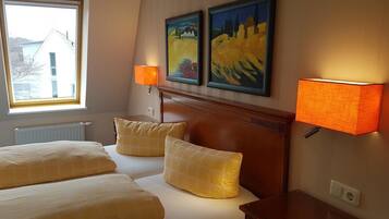 Quarto de hotel com duas camas, cabeceira de madeira, janela com vista e dois quadros na parede.