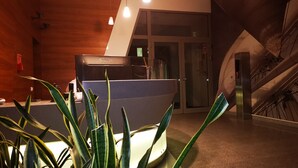 Uma área de recepção com design moderno, destacando um balcão de atendimento, uma grande planta em vaso e uma escadaria ao fundo.