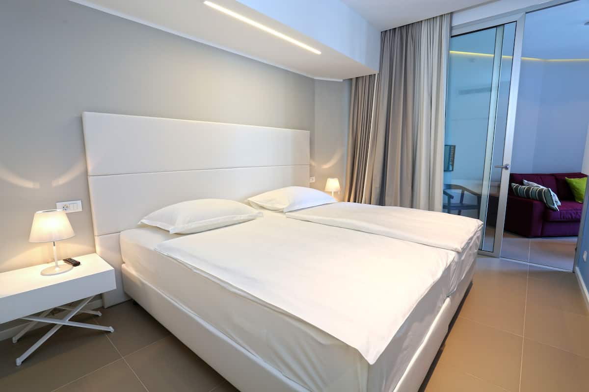 Quarto de hotel moderno com uma cama grande, uma mesa de cabeceira com abajur e vista para uma varanda com sofá roxo.