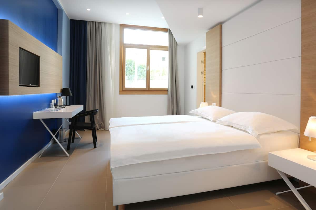 Quarto de hotel moderno com uma cama grande, uma escrivaninha com cadeira, uma TV na parede e uma janela com cortinas.