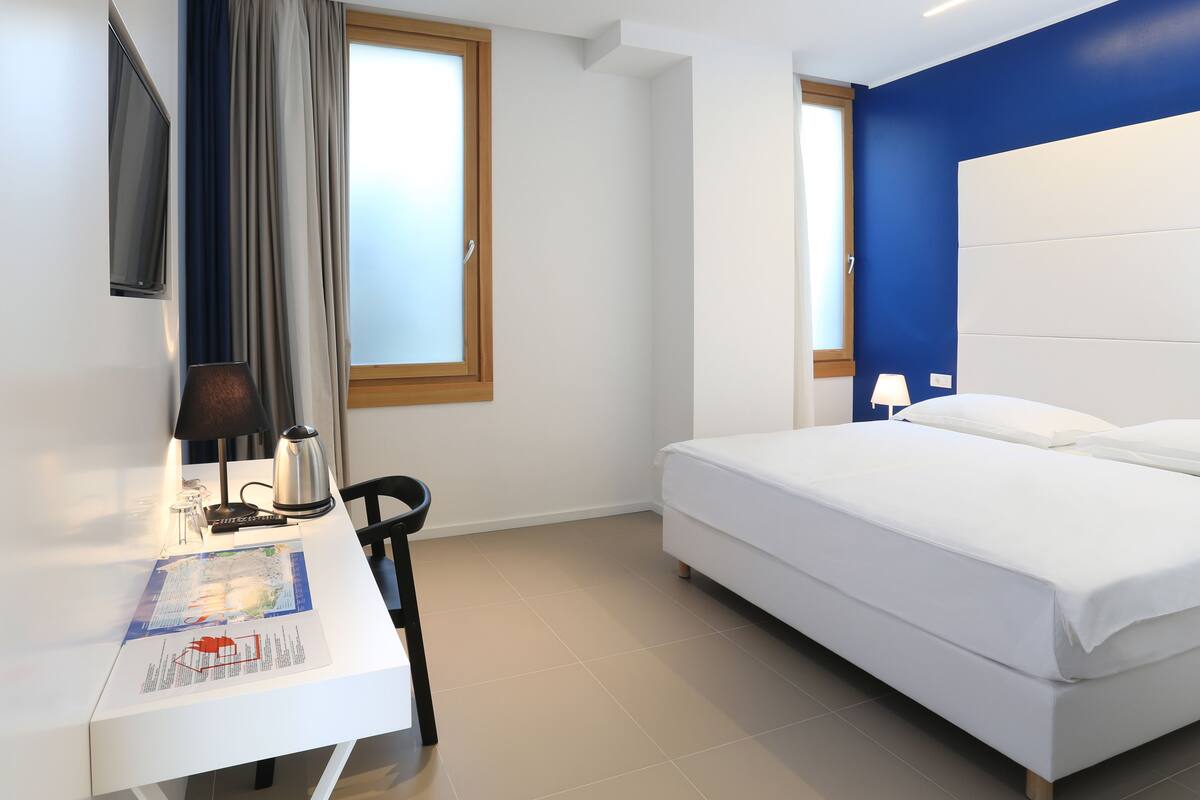 Quarto de hotel com cama, escrivaninha com luminária, cafeteira, cadeira e janela com cortinas.
