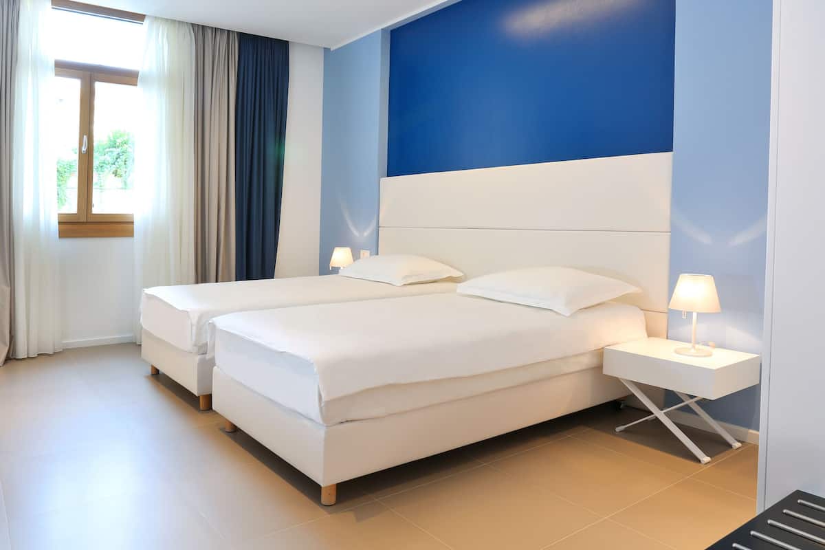 Quarto de hotel com duas camas, uma parede com detalhes em azul e uma janela com cortinas.