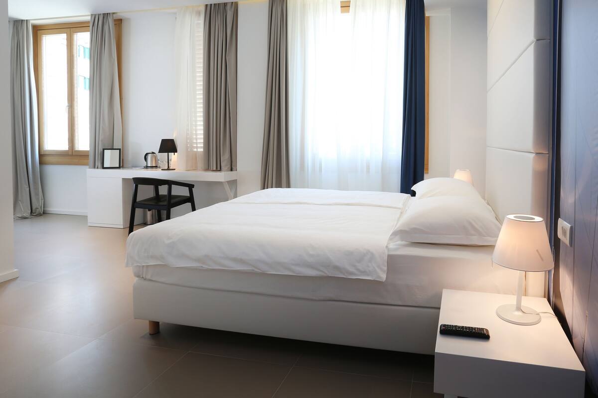 Quarto de hotel moderno com uma cama grande, uma escrivaninha, uma cadeira e uma mesinha de cabeceira com luminária e controle remoto.