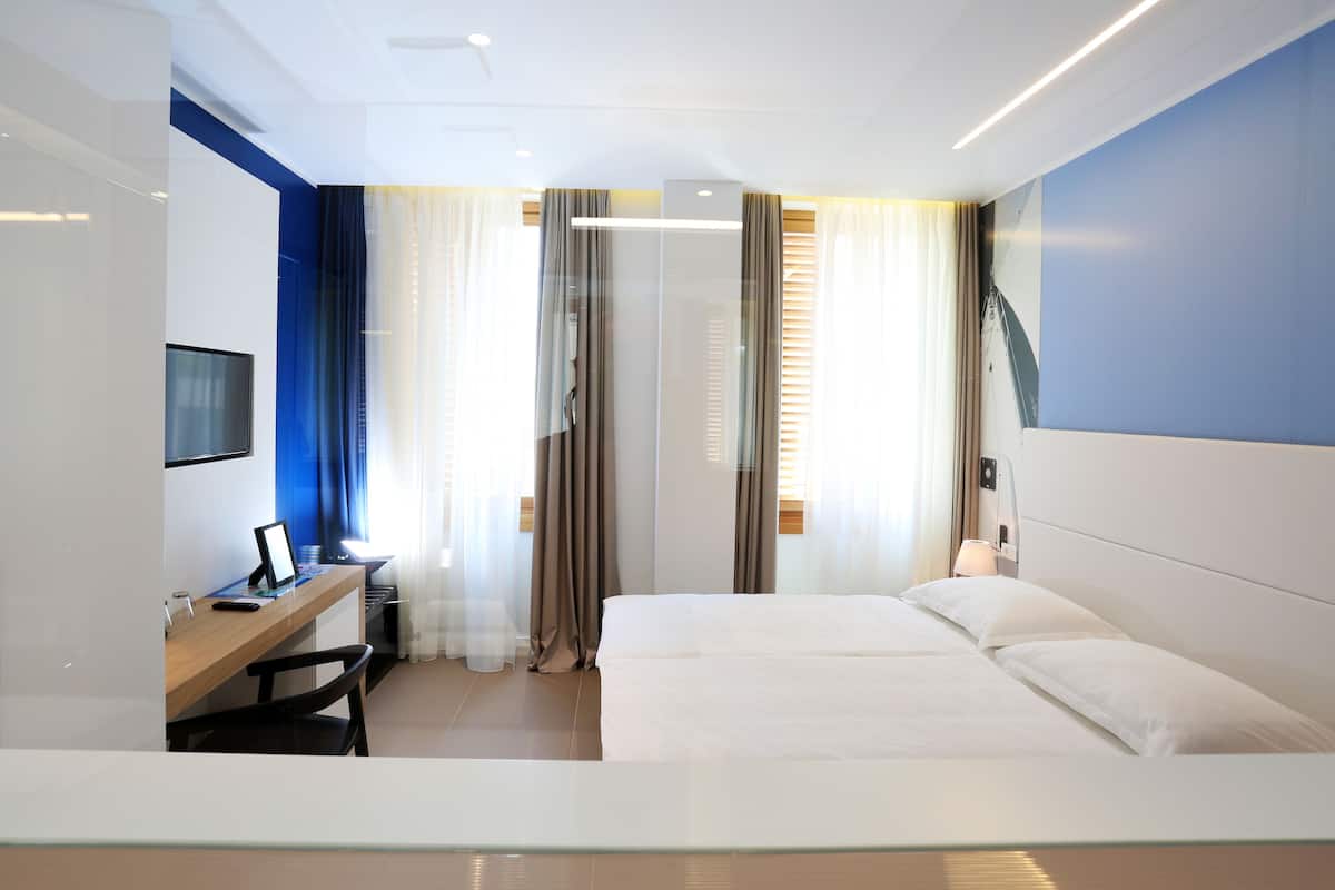 Quarto de hotel moderno com uma cama grande, uma escrivaninha com cadeira, uma televisão e uma janela com cortinas.