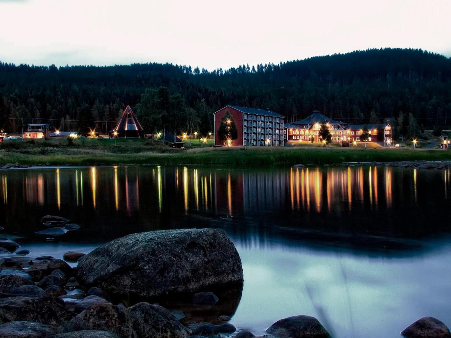 Hotel à beira do lago, com o reflexo dos edifícios na água, rodeado por uma área arborizada.