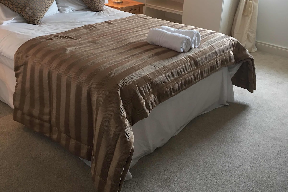 Cama bem arrumada com um cobertor listrado, duas almofadas estampadas e uma toalha dobrada ao lado. Há um abajur, um guarda-roupa branco e uma janela com cortinas.
