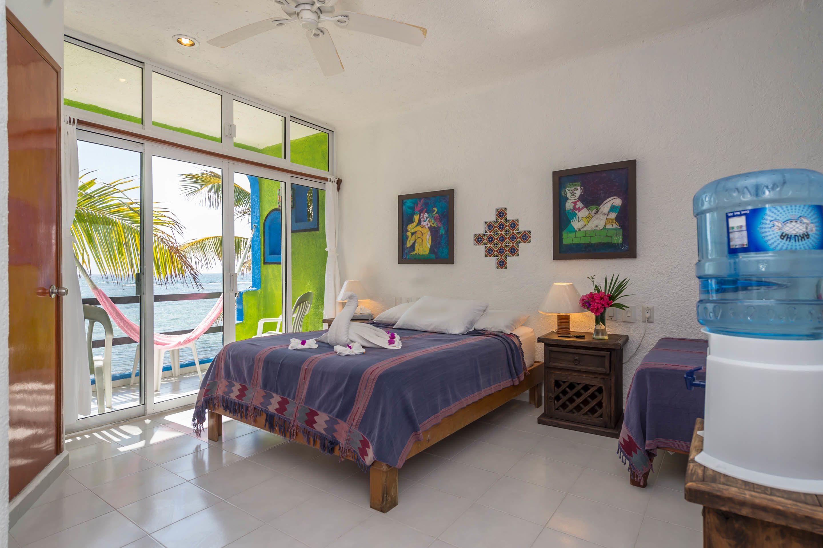 Quarto com cama, mesa de cabeceira, dispensador de água e vista para o oceano através de uma porta de vidro.