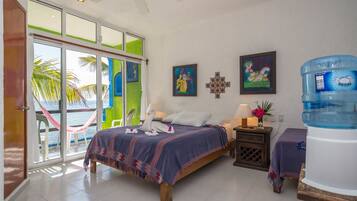 Quarto com cama, mesa de cabeceira, dispensador de água e vista para o oceano através de uma porta de vidro.