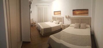 Quarto de hotel com duas camas, uma cadeira, uma mesinha de cabeceira e duas luminárias fixadas na parede.