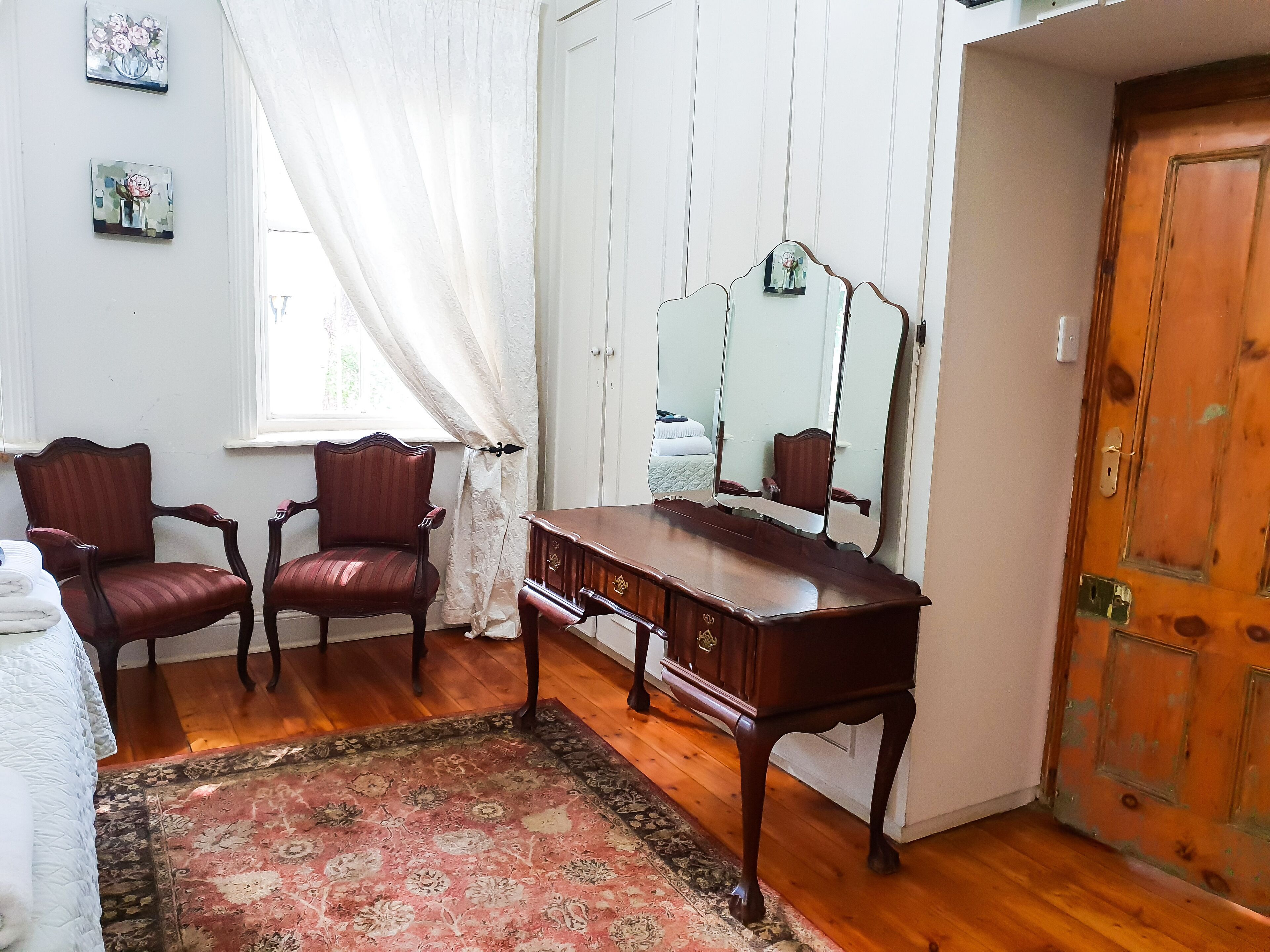 Quarto com duas cadeiras de madeira, cômoda de madeira, espelho, tapete estampado e porta de madeira.