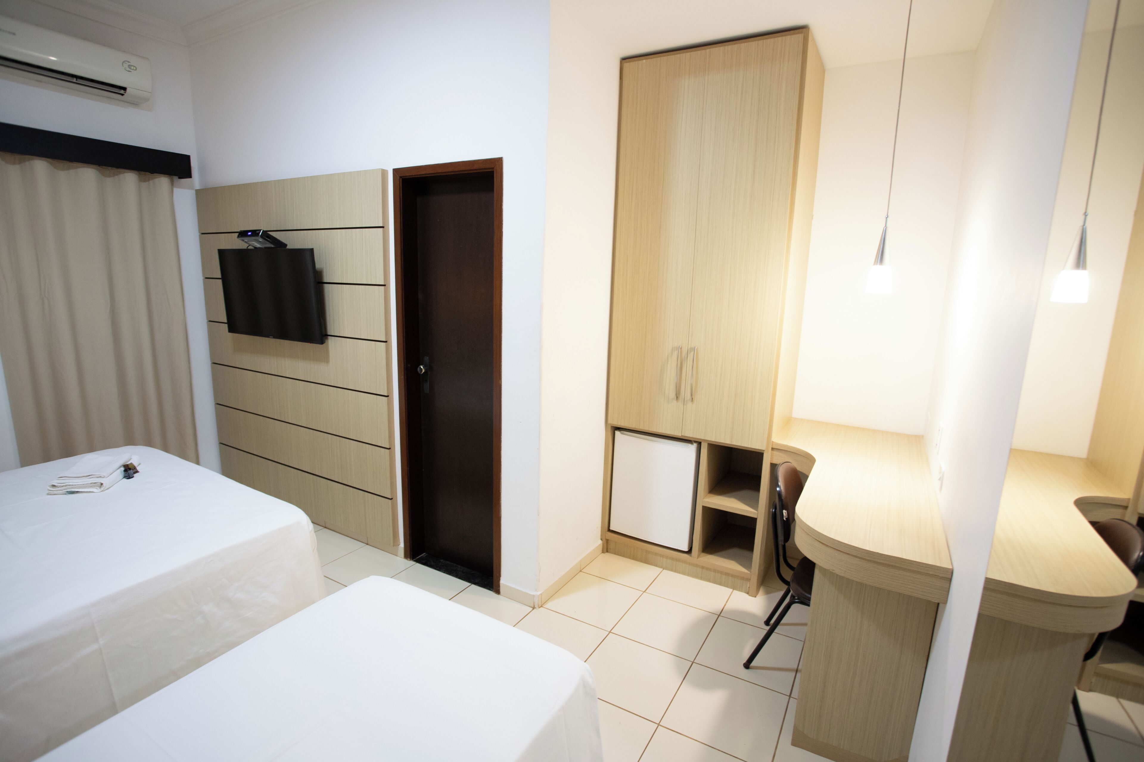Quarto de hotel com duas camas, um guarda-roupa, uma escrivaninha e uma cadeira.