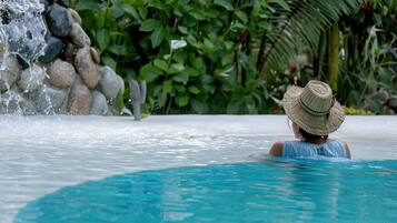 Uma pessoa em uma piscina com cascata e vegetação exuberante ao fundo.