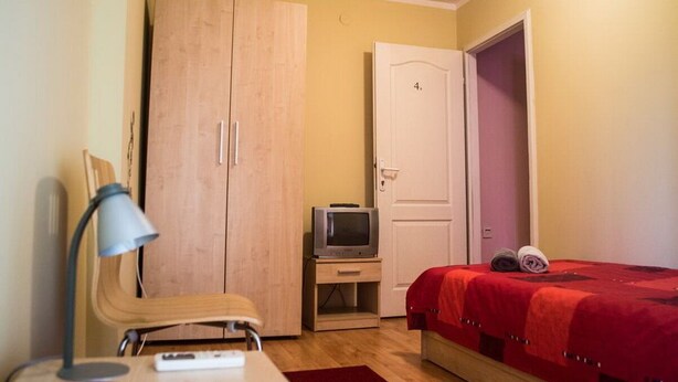 Quarto com cama, televisão sobre suporte, guarda-roupa, secretária com luminária e uma cadeira.