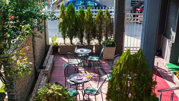Um pátio externo com mesas e cadeiras, cercado por plantas em vasos e um chão de azulejos vermelhos.