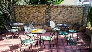 Uma área de pátio ao ar livre com mesas e cadeiras redondas, uma parede de tijolos e piso de azulejos.