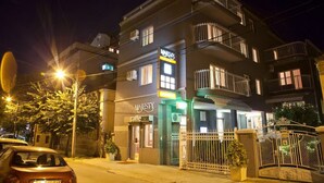 Um edifício de vários andares com um café chamado 'MAJESTY' no andar térreo, uma entrada visível e um portão com uma pequena árvore na frente.