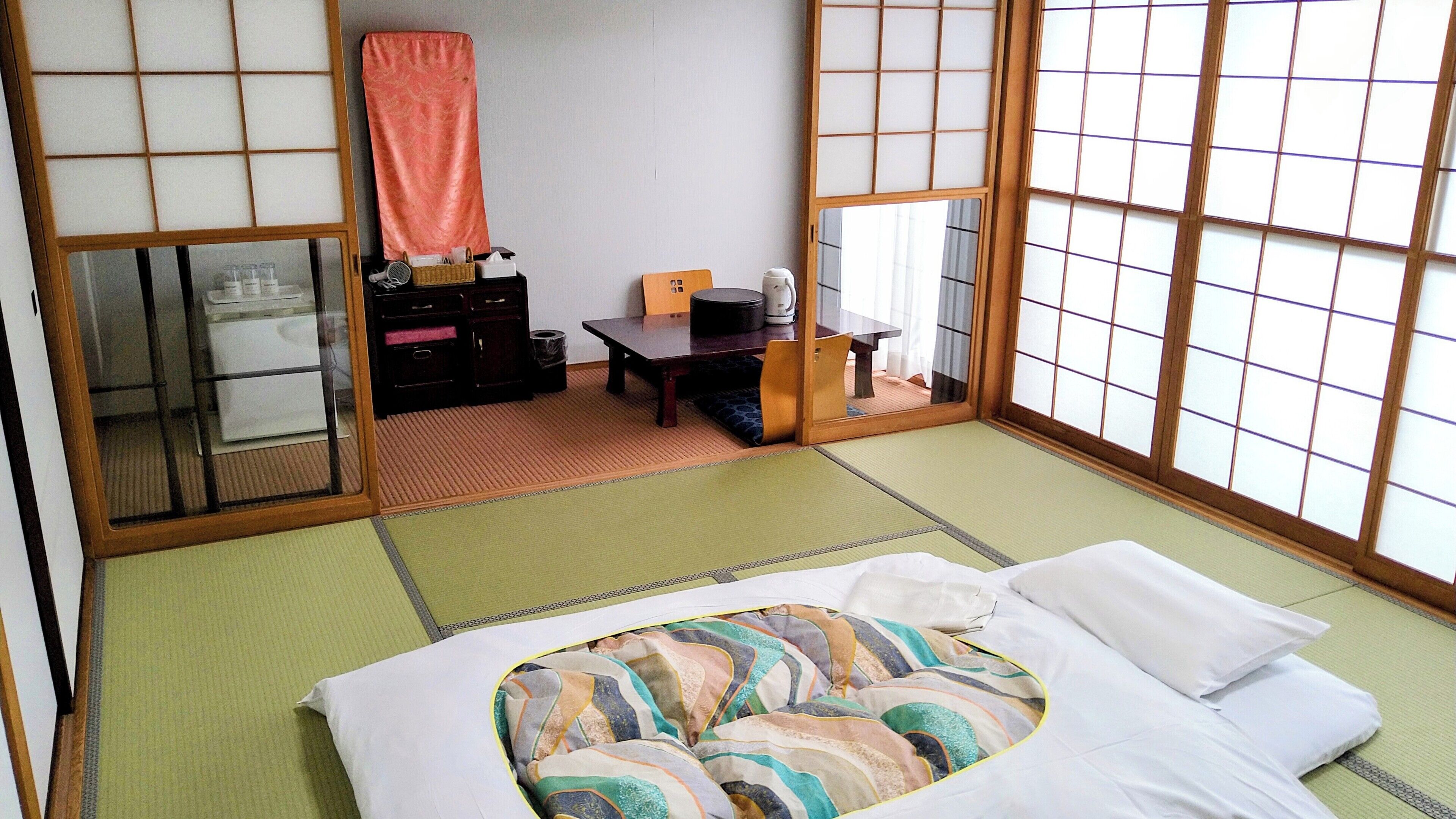 Um quarto tradicional japonês com tatames, uma cama com manta colorida, uma escrivaninha com chaleira e portas corrediças.