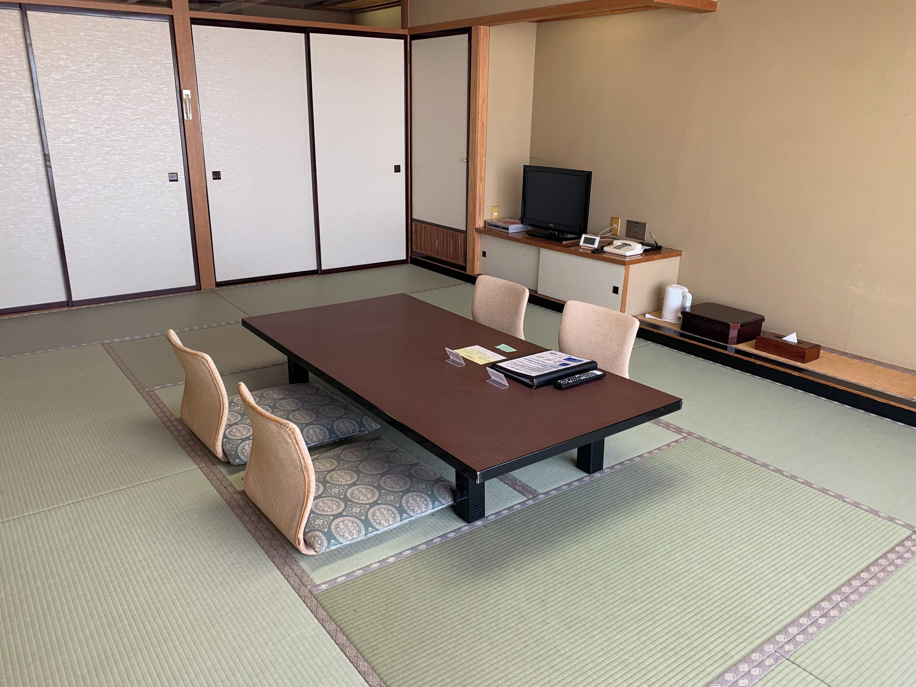Um quarto tradicional japonês com piso de tatame, uma mesa baixa com almofadas, uma televisão e um pequeno armário com um telefone e outros objetos.