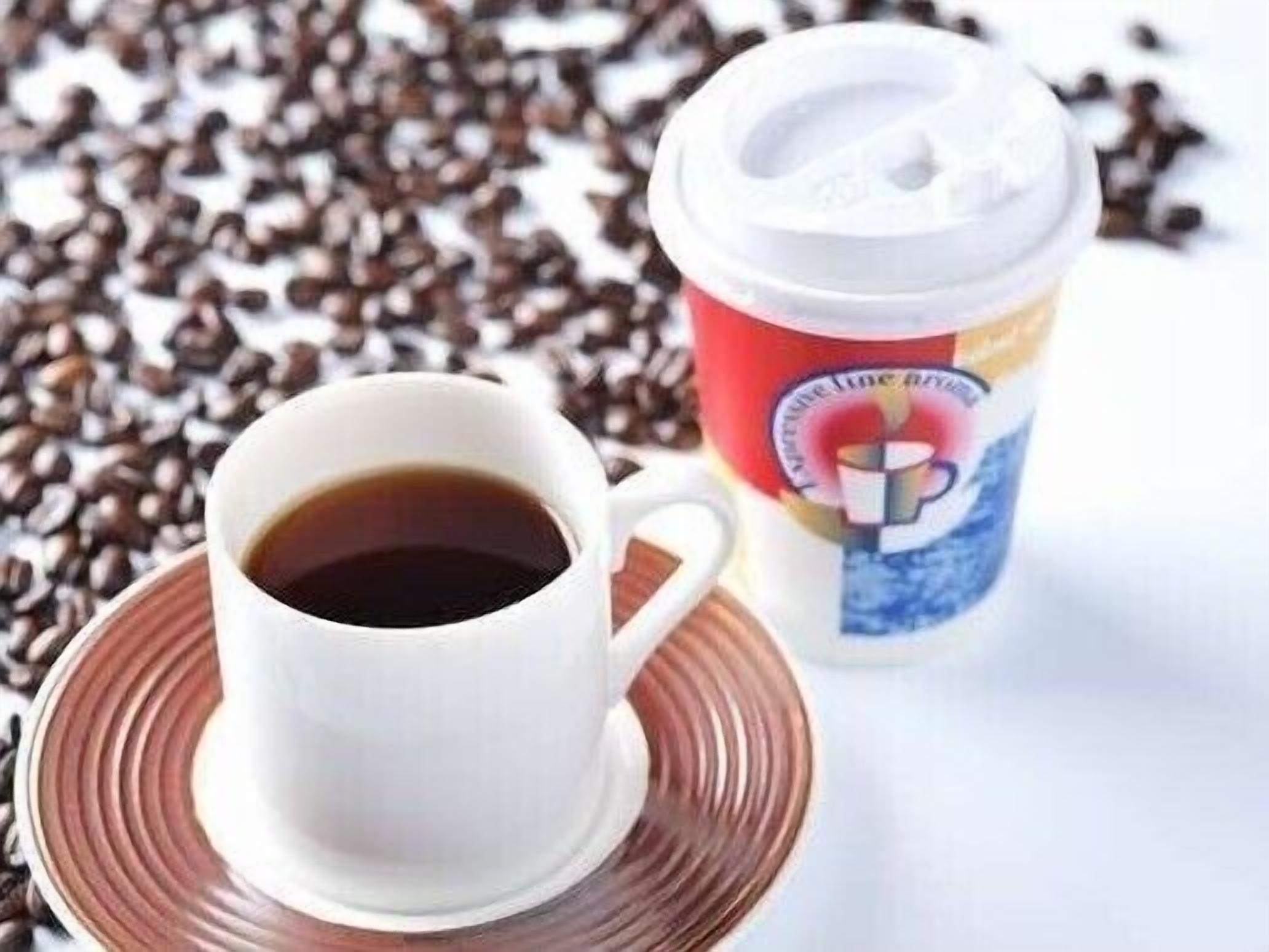 Uma chávena de café branca num pires, uma chávena de café vermelha e branca com tampa, e grãos de café espalhados.
