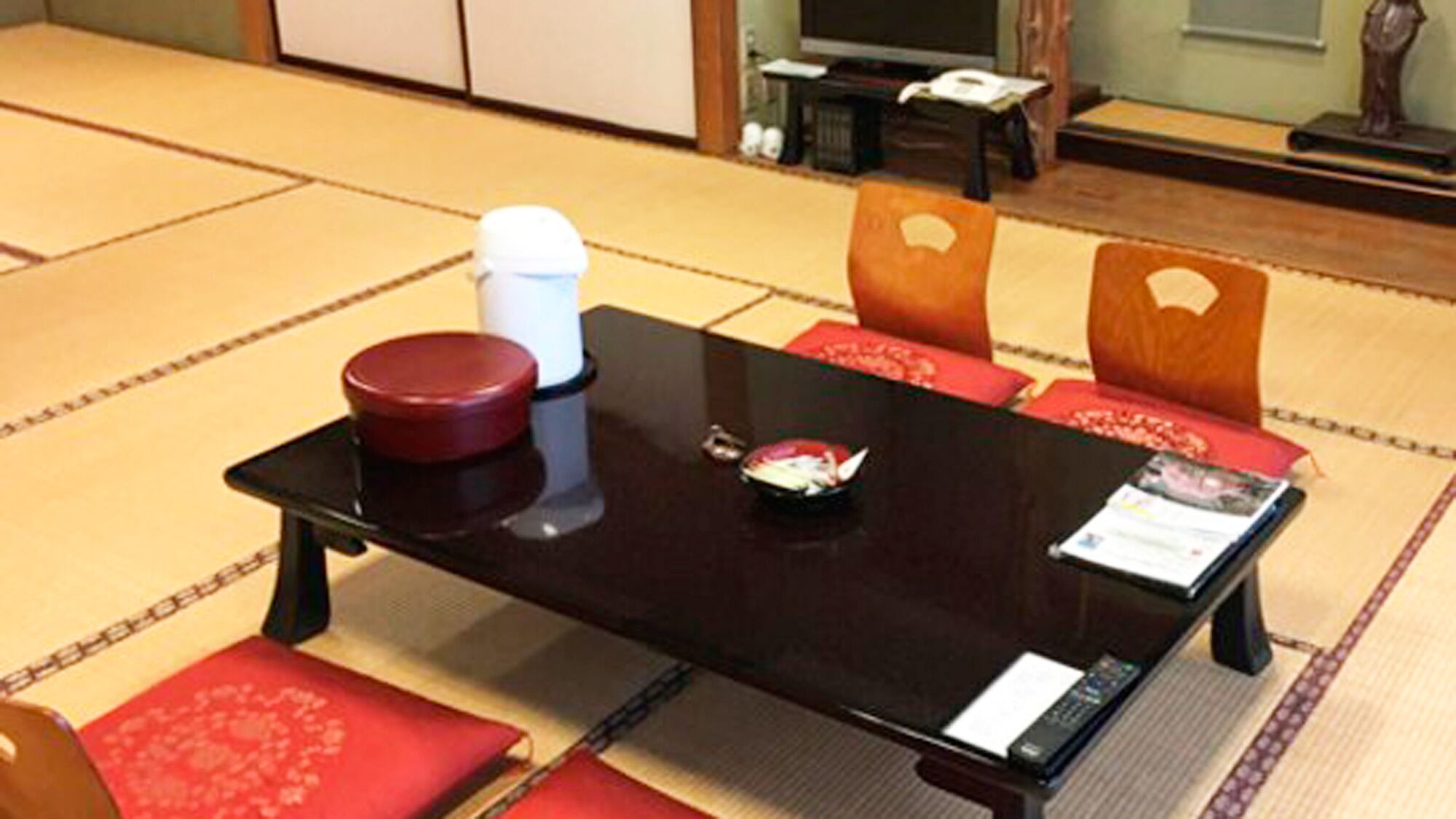 Um quarto tradicional com esteiras de tatame, uma mesa baixa, duas almofadas vermelhas, um recipiente branco e um controle remoto sobre a mesa.
