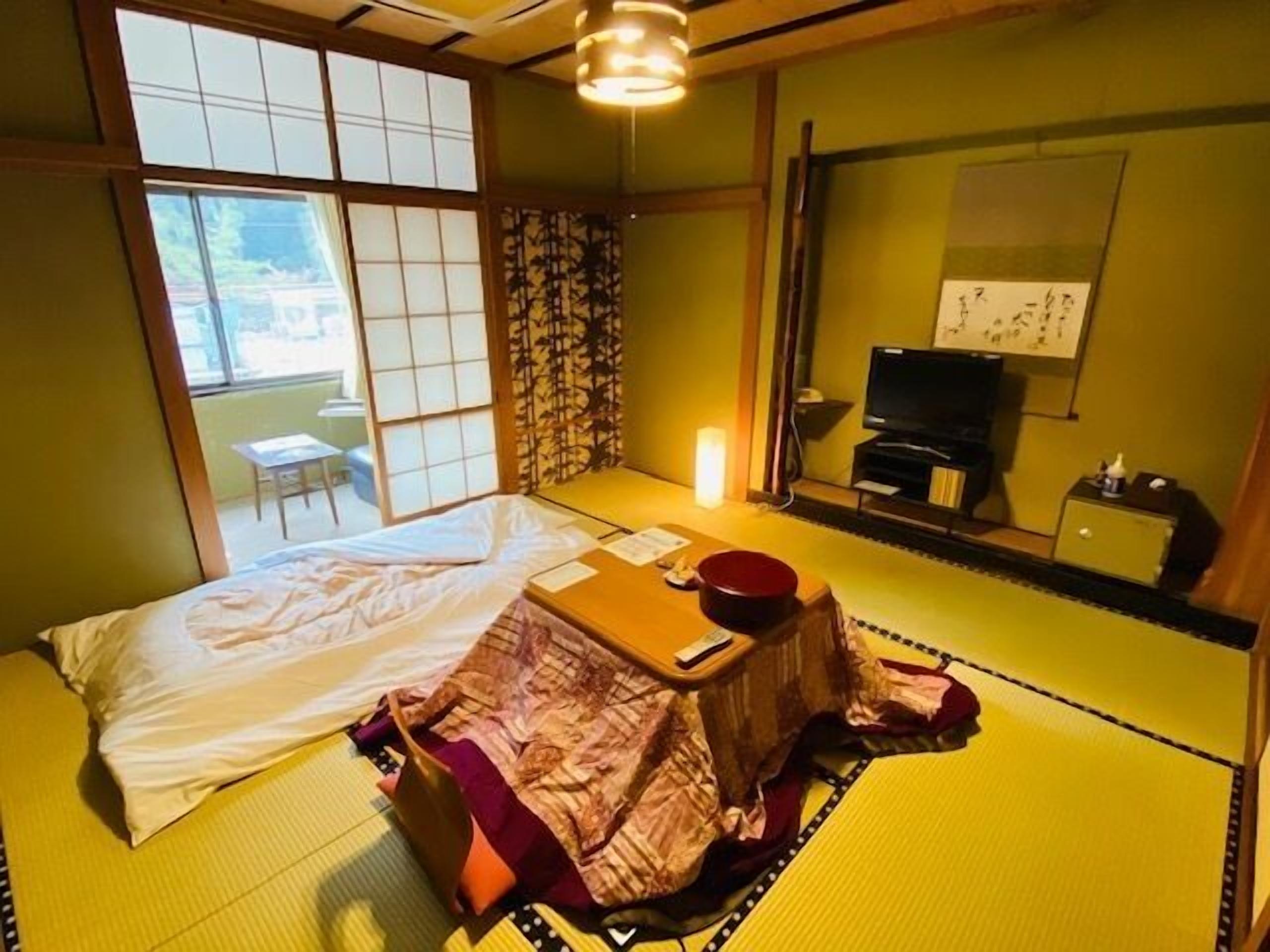 Quarto tradicional japonês com tatames, uma cama com roupa de cama branca, uma pequena mesa com uma tigela vermelha, uma televisão e uma porta deslizante que dá para uma varanda.