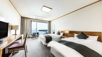 Quarto de hotel com duas camas, uma escrivaninha com cadeira, televisão e vista para o mar.