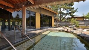 Uma área de onsen tradicional japonesa ao ar livre, com pérgola de madeira, degraus de pedra e uma pequena piscina.