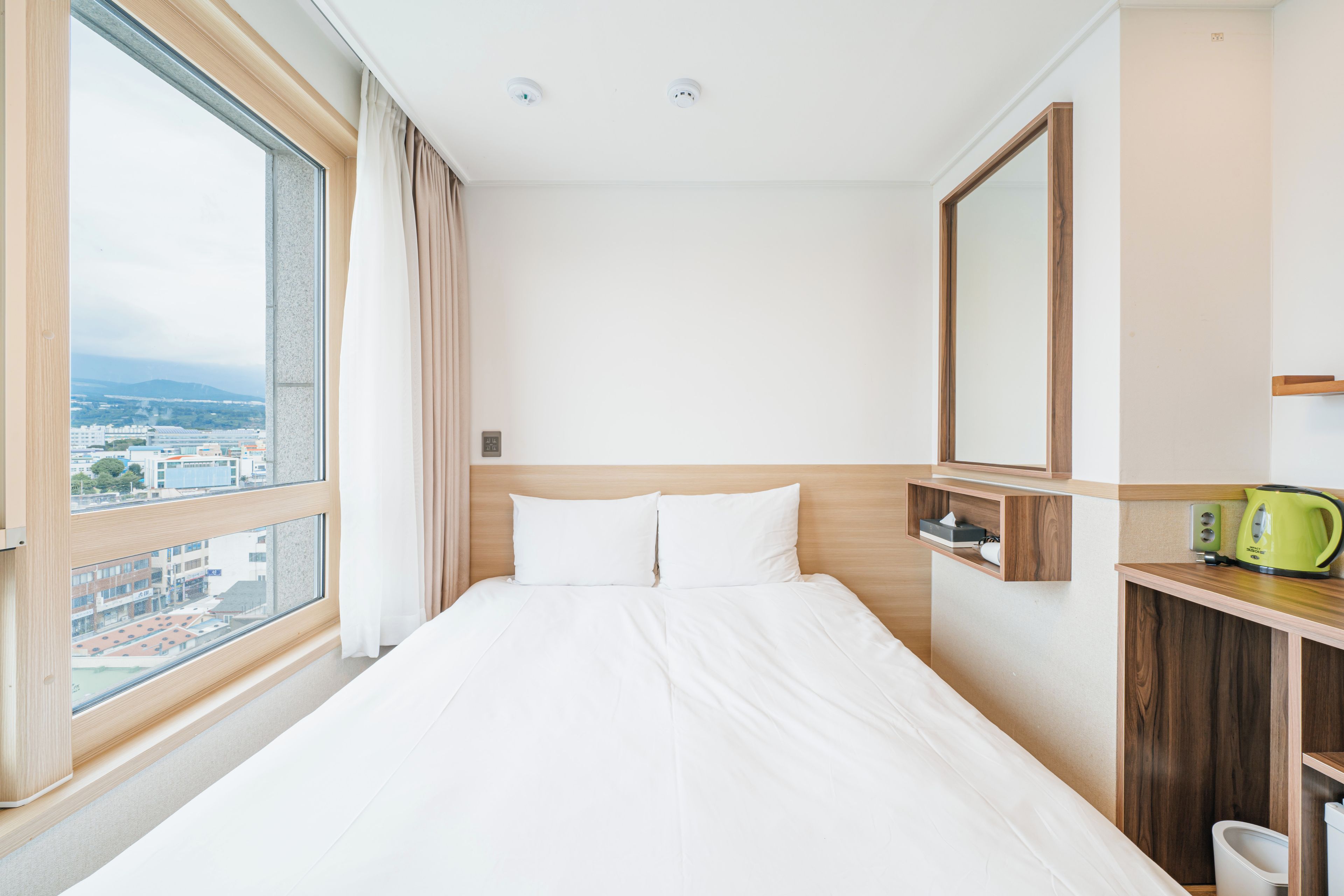 Quarto de hotel com uma janela grande, cama com roupa branca, cabeceira de madeira, pequena estante com chaleira e um espelho na parede.