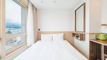 Quarto de hotel com uma janela grande, cama com roupa branca, cabeceira de madeira, pequena estante com chaleira e um espelho na parede.