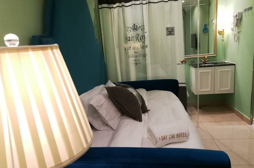 Quarto de hotel com cama, mesa de cabeceira, espelho, abajur e banheiro com pia e vaso sanitário.