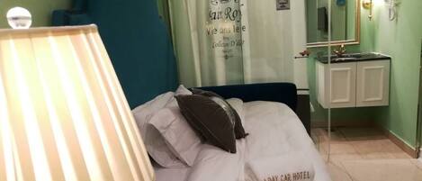 Quarto de hotel com cama, mesa de cabeceira, espelho, abajur e banheiro com pia e vaso sanitário.
