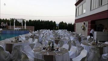 Uma área de banquetes ao ar livre com mesas redondas preparadas para refeições, cercada por capas brancas nas cadeiras.