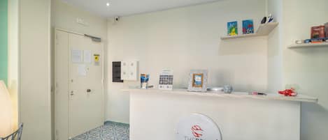 Uma área de recepção com balcão exibindo um logotipo, um extintor de incêndio e uma mangueira. Há prateleiras com livros e objetos decorativos, e um piso estampado.