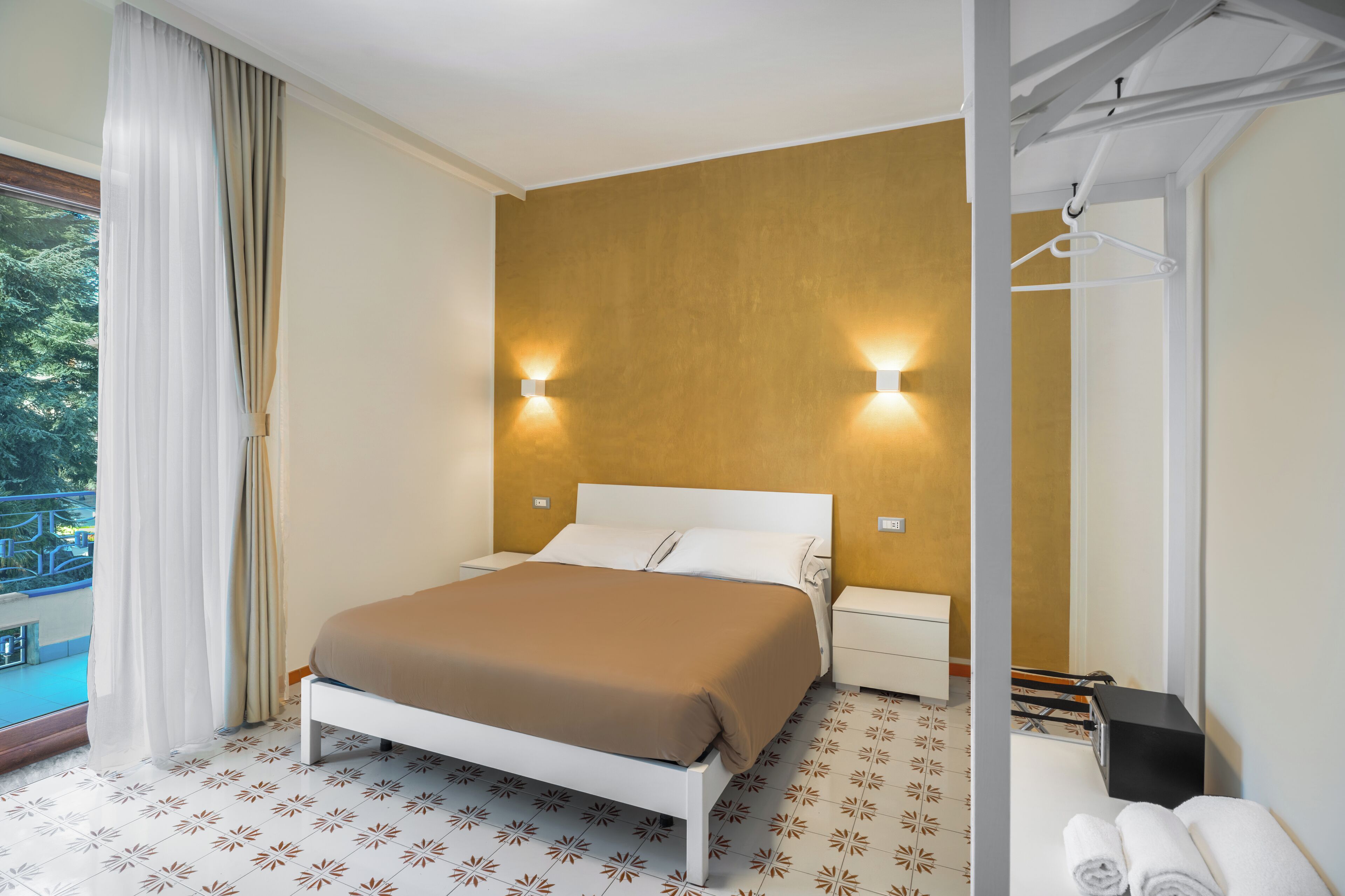 Quarto de hotel com uma cama, duas mesas de cabeceira, uma luminária e uma janela com cortinas, oferecendo vista para a piscina.