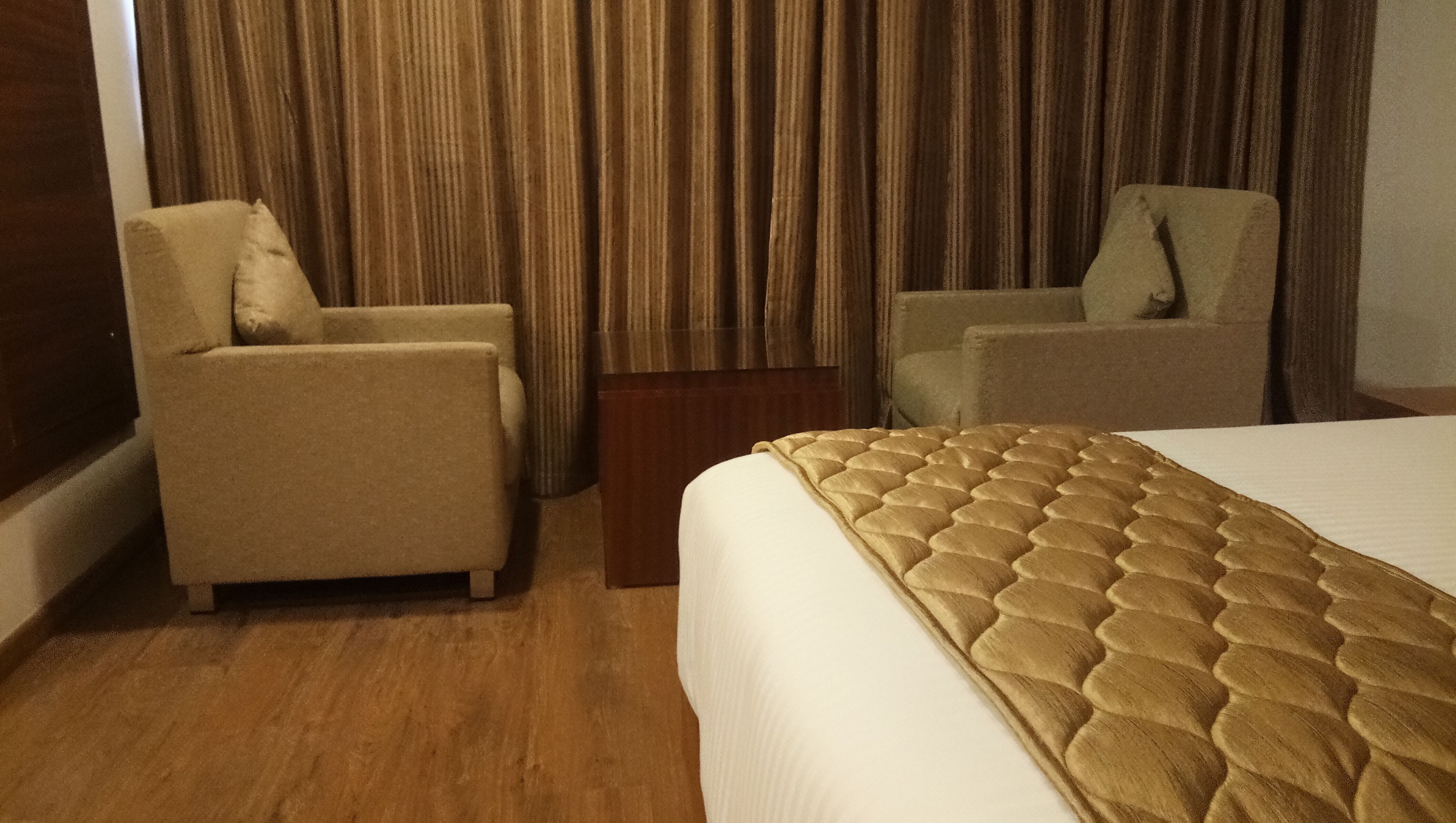 Quarto de hotel com cama, duas poltronas, mesa de cabeceira de madeira e cortina listrada.