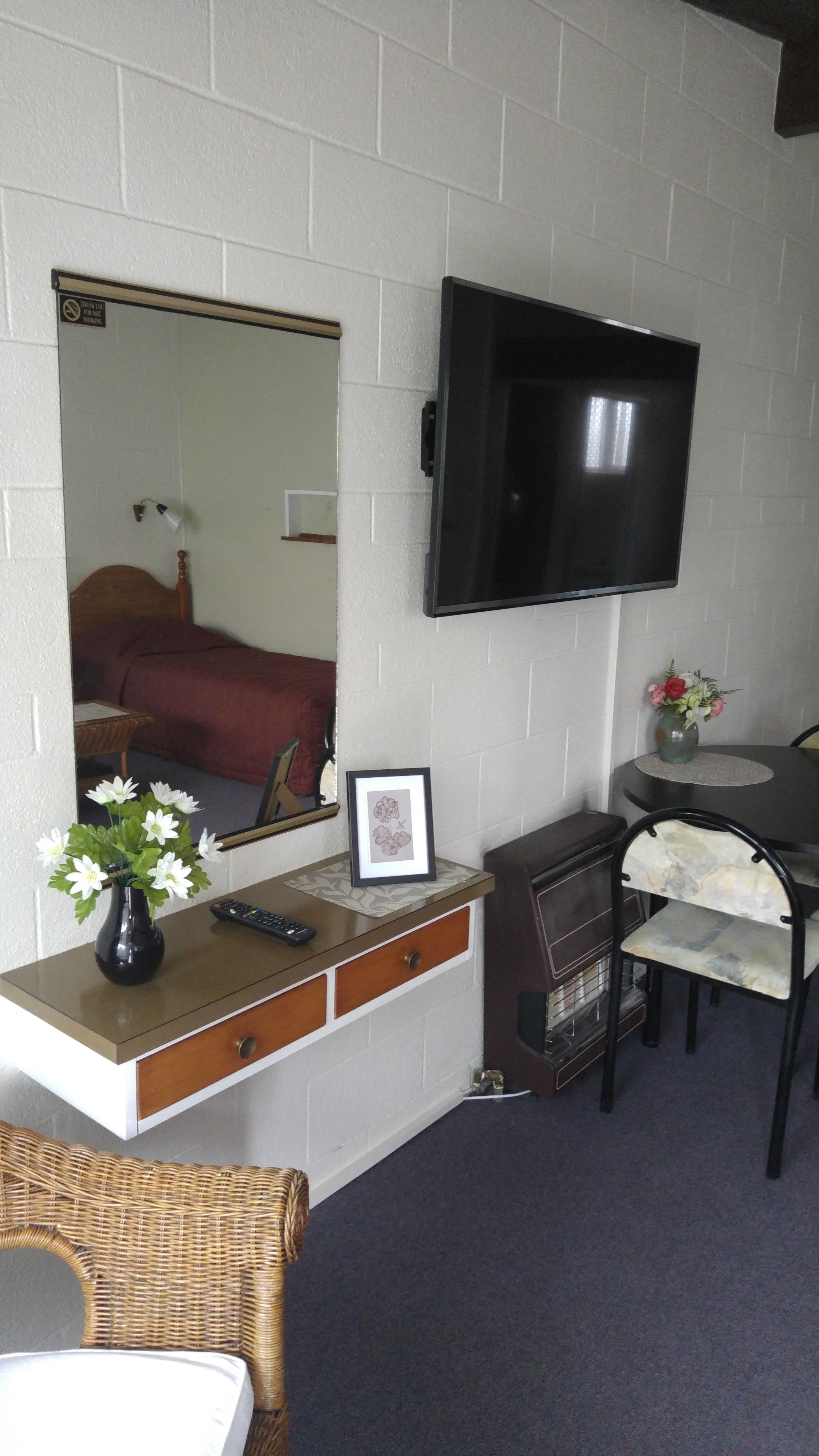 Quarto de hotel com cama, televisão na parede, mesinha com um vaso de flores, quadro e cadeira de vime.