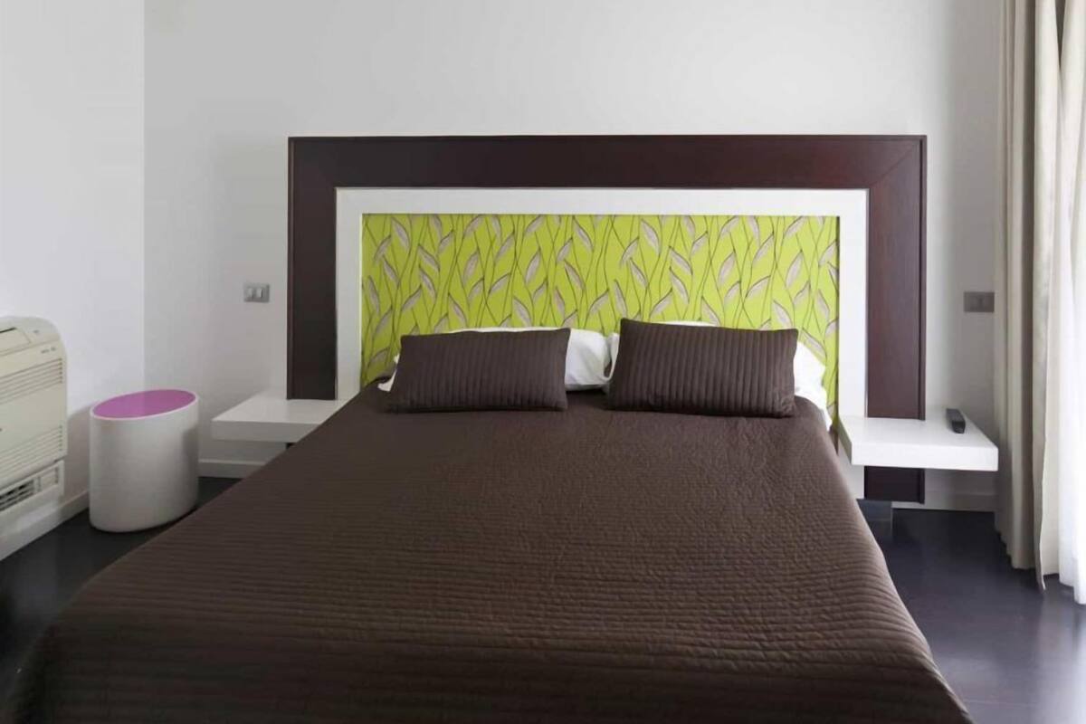 Quarto de hotel moderno com uma cama grande, cabeceira com padrão de folha, mesa de cabeceira e luminária pendente.