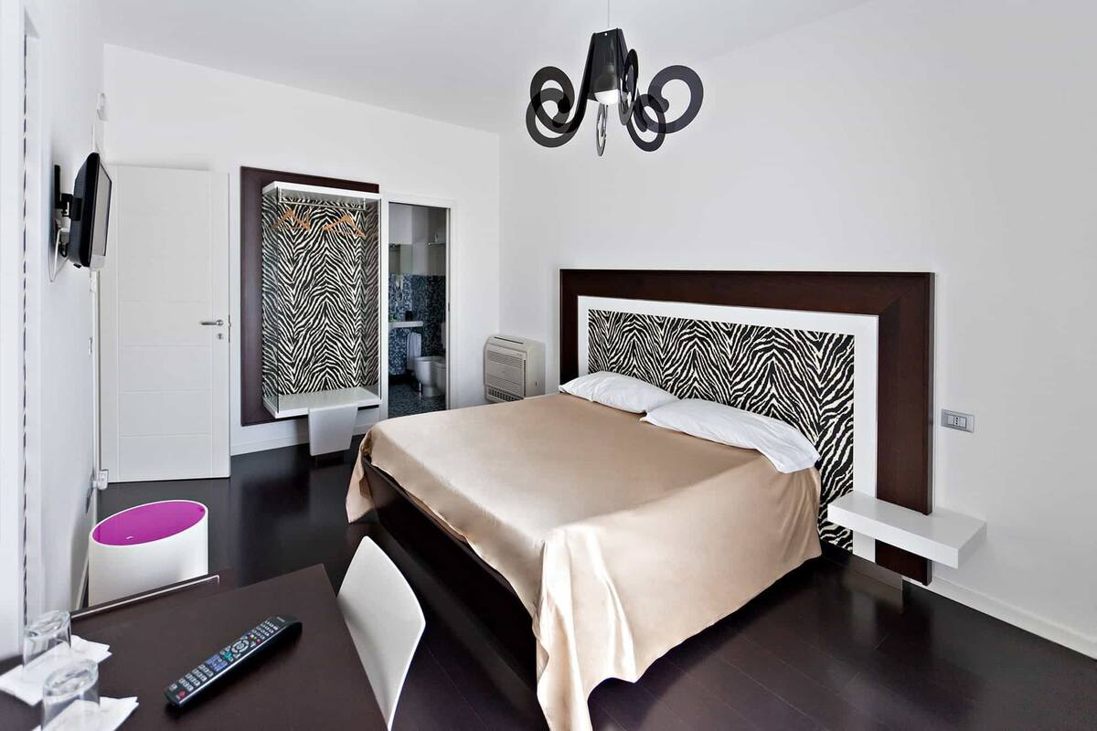 Quarto de hotel moderno com uma cama grande, uma escrivaninha com controle remoto e um banheiro visível através de uma porta aberta.