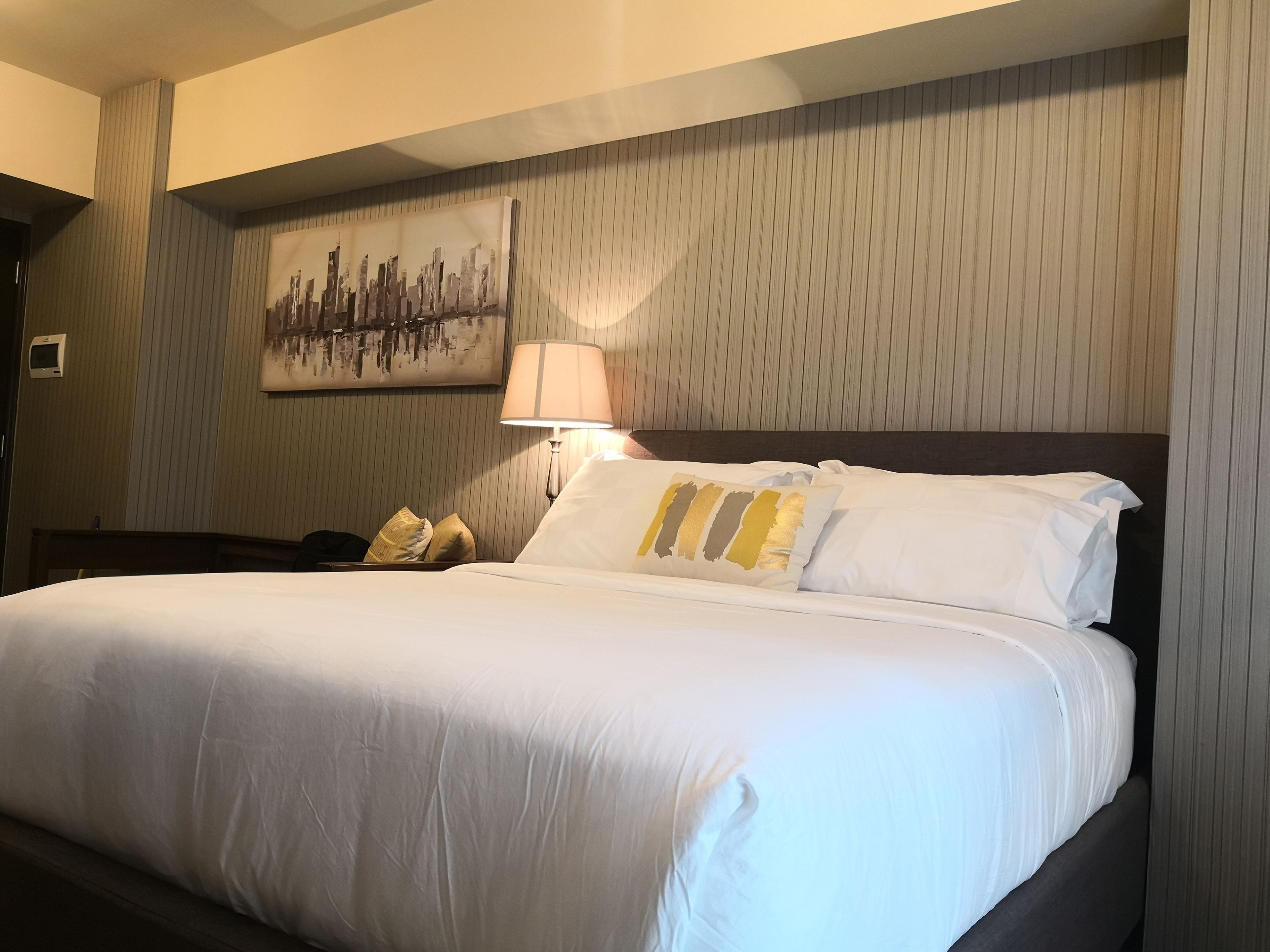 Quarto de hotel com uma cama grande, uma mesa de cabeceira, um abajur e uma pintura com vista para a cidade na parede.