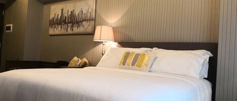 Quarto de hotel com uma cama grande, uma mesa de cabeceira, um abajur e uma pintura com vista para a cidade na parede.