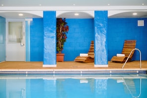 Piscina com paredes e colunas azuis, duas espreguiçadeiras de madeira e um vaso com planta.